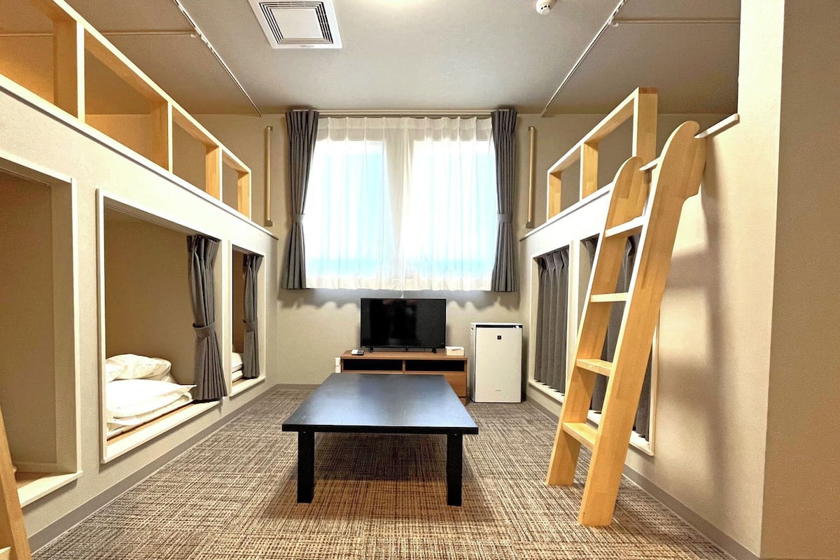Quarto com beliches, televisão, uma mesinha e janela com cortinas.