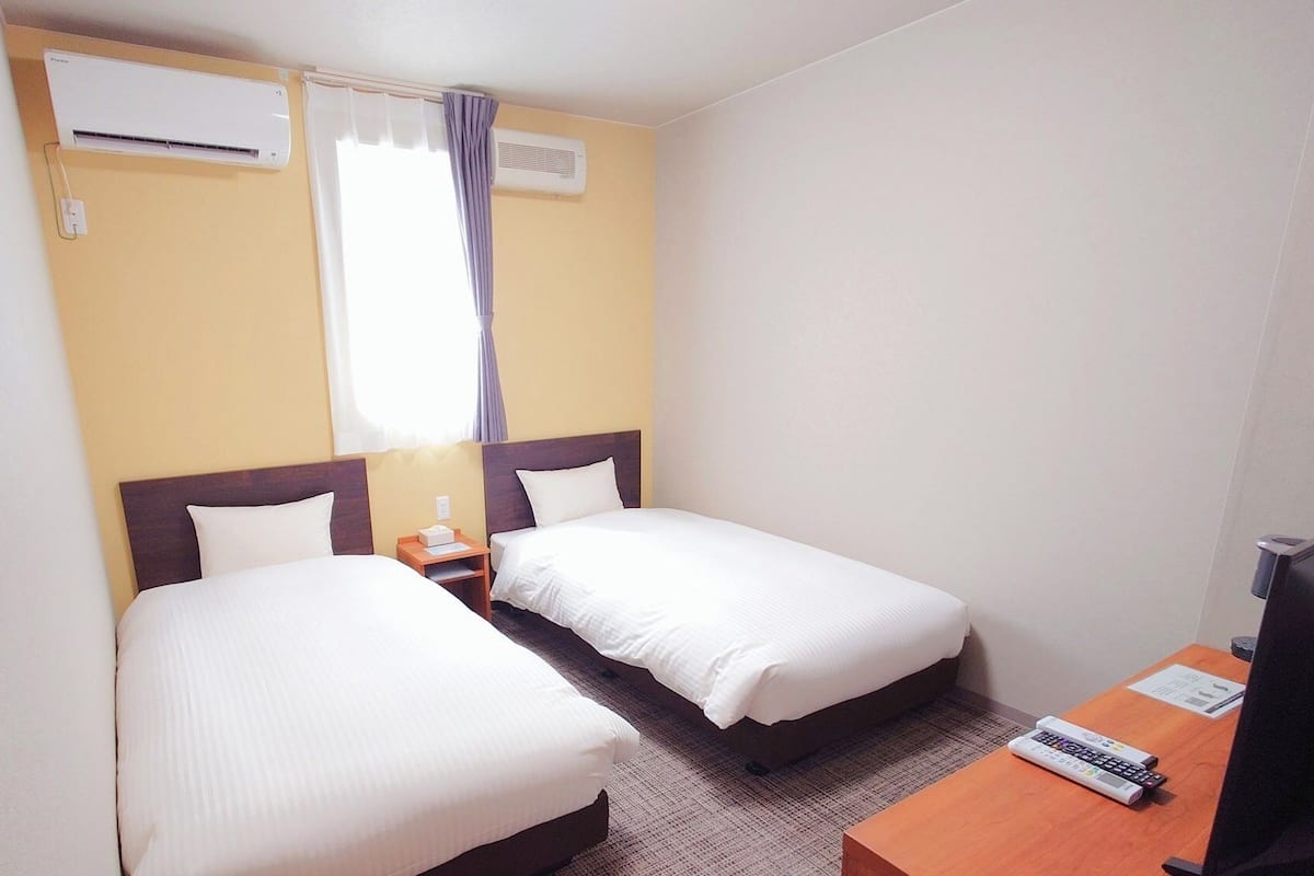 Quarto de hotel com duas camas de solteiro, uma mesinha de cabeceira de madeira, uma escrivaninha com televisão e dois aparelhos de ar condicionado na parede.