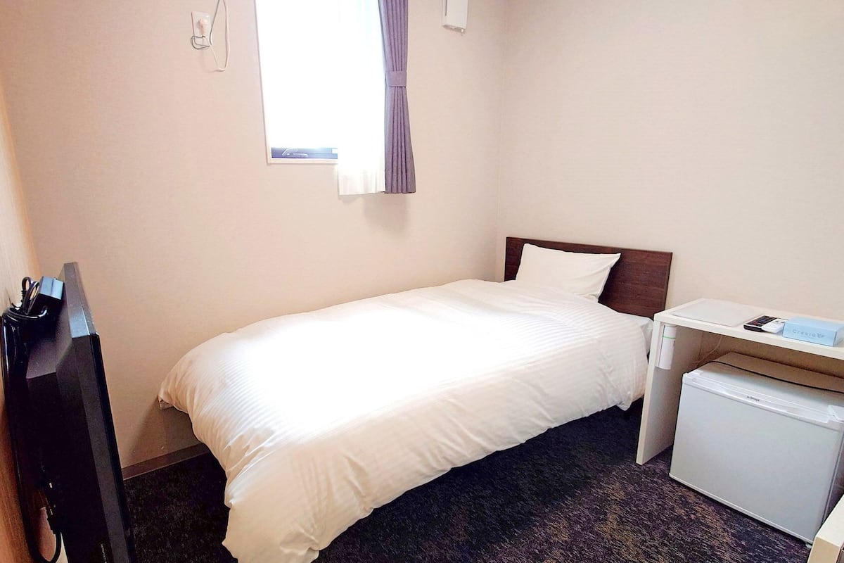 Quarto de hotel com cama, escrivaninha, pequeno frigorífico e janela com cortinas.