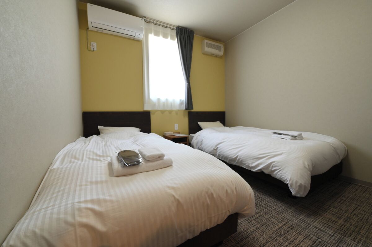 Quarto de hotel com duas camas, janela com cortinas, aparelho de ar condicionado e piso coberto por carpete.