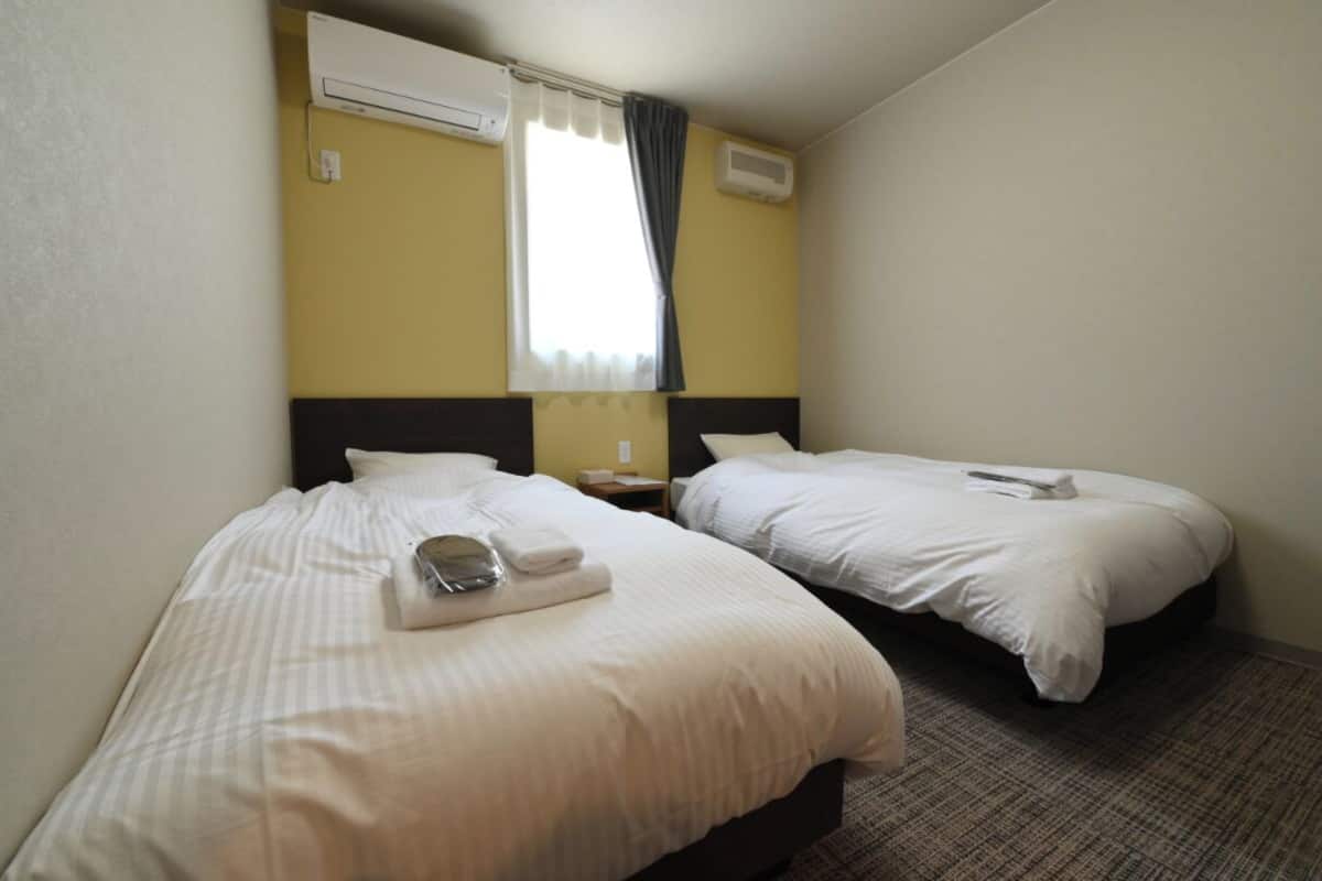 Quarto de hotel com duas camas, janela com cortinas, aparelho de ar condicionado e piso coberto por carpete.