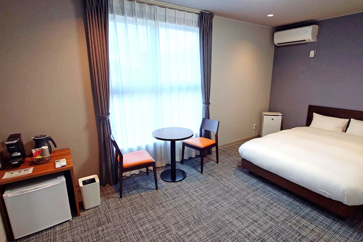 Quarto de hotel com cama, escrivaninha com cafeteira, cadeira, mesinha e ar condicionado.
