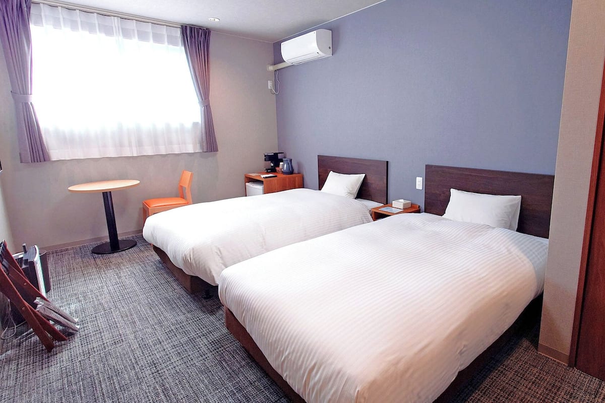 Quarto de hotel com duas camas, uma mesinha, uma cadeira, uma escrivaninha e um ar condicionado.