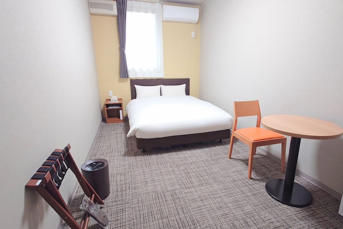 Quarto de hotel com uma cama, uma cadeira, uma mesa, um cesto de lixo e um aparelho de ar condicionado.
