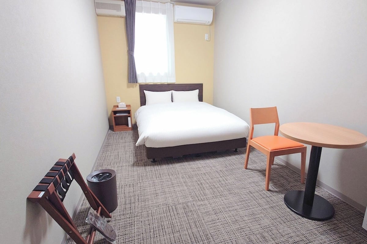 Quarto de hotel pequeno com uma cama de solteiro, uma cadeira, uma mesinha, um cesto de lixo e um aparelho de ar condicionado.