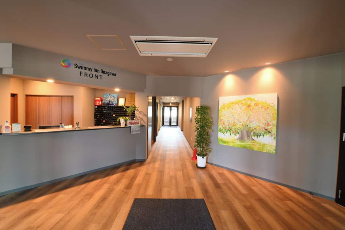 Uma área de recepção com balcão, placa com 'Swimmy Inn Onagawa FRONT', uma pintura de árvore e um corredor que leva a outro quarto.