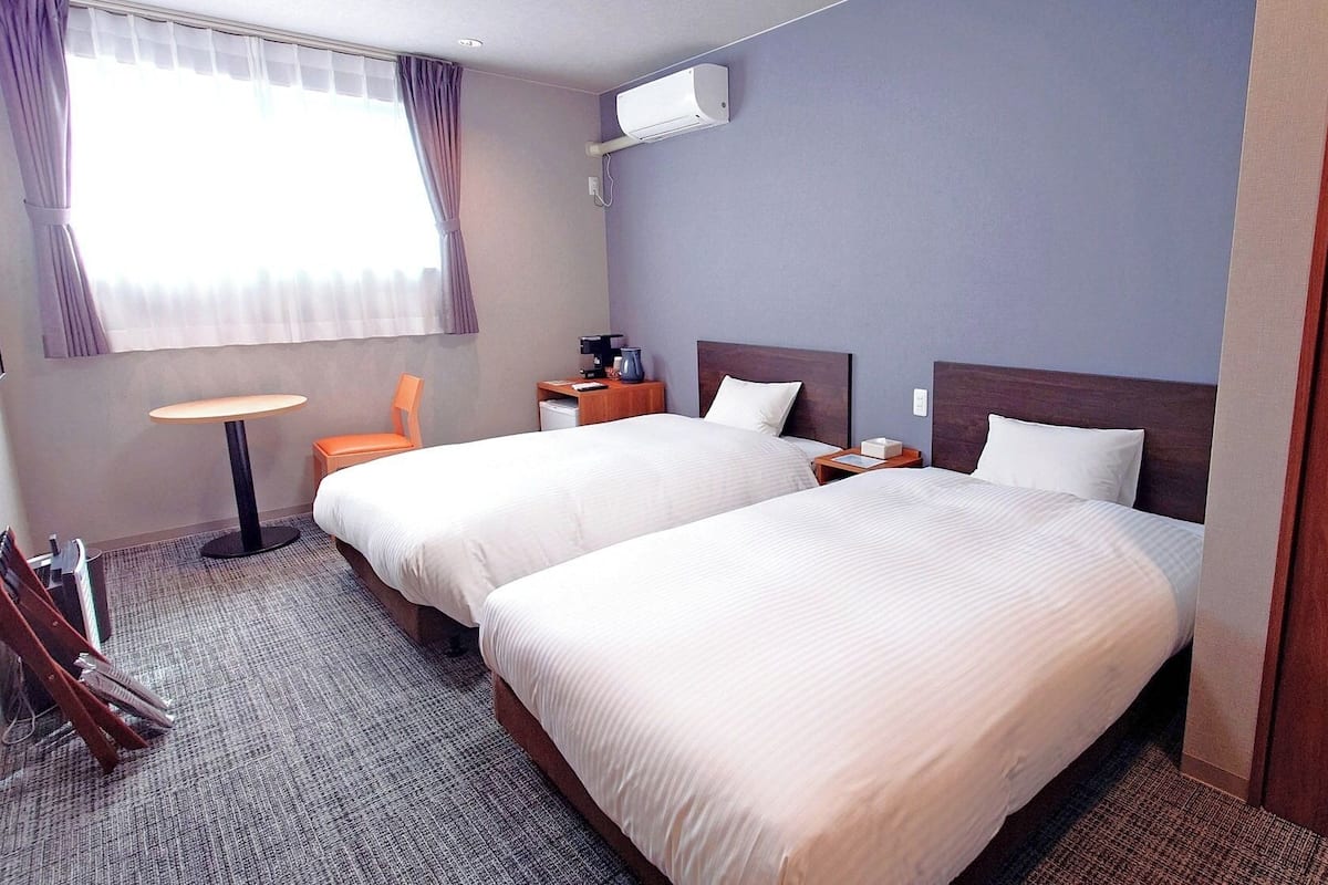 Quarto de hotel com duas camas, uma mesinha, uma cadeira, uma escrivaninha com abajur e um aparelho de ar condicionado.