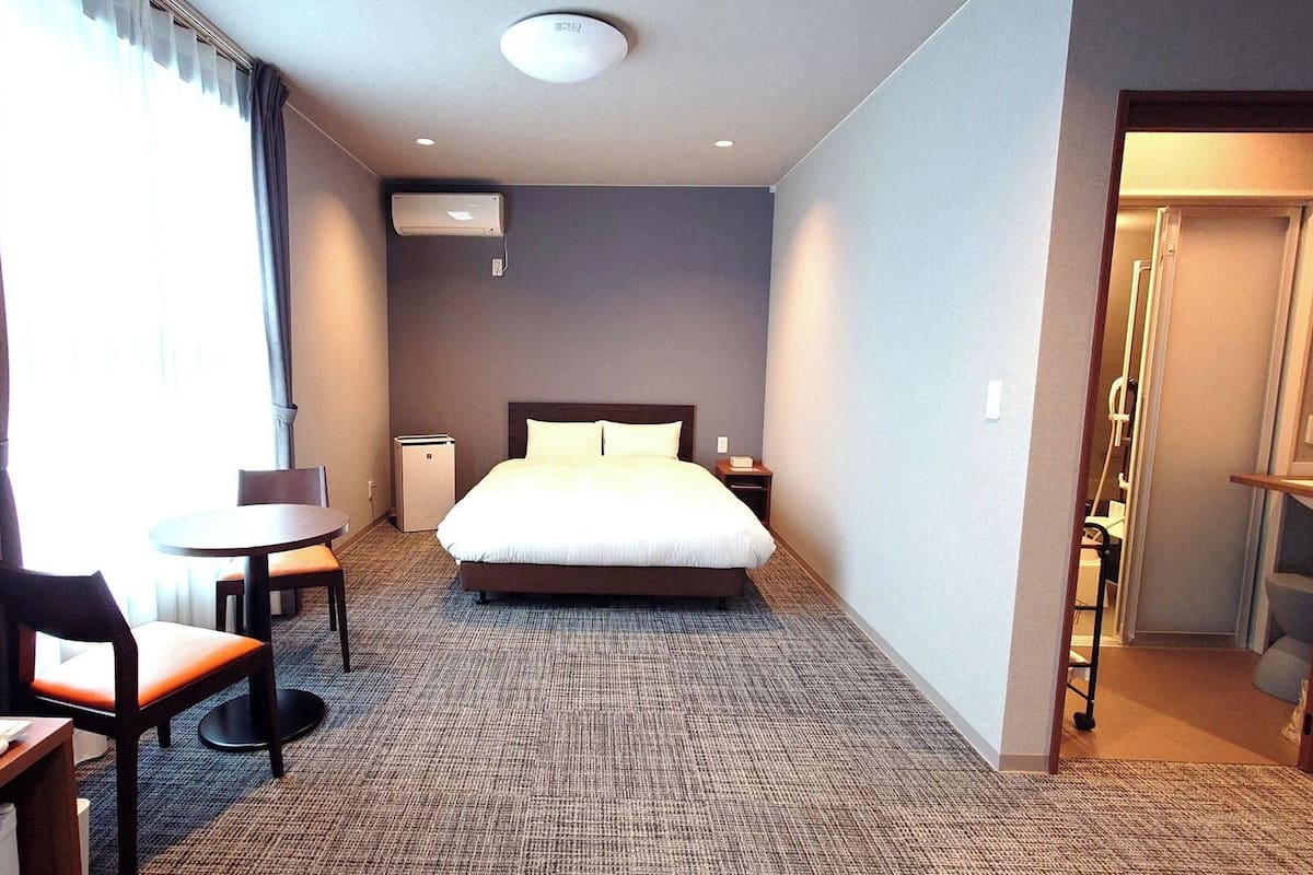 Quarto de hotel com cama, escrivaninha, cadeira, mesa pequena e janela com cortinas.
