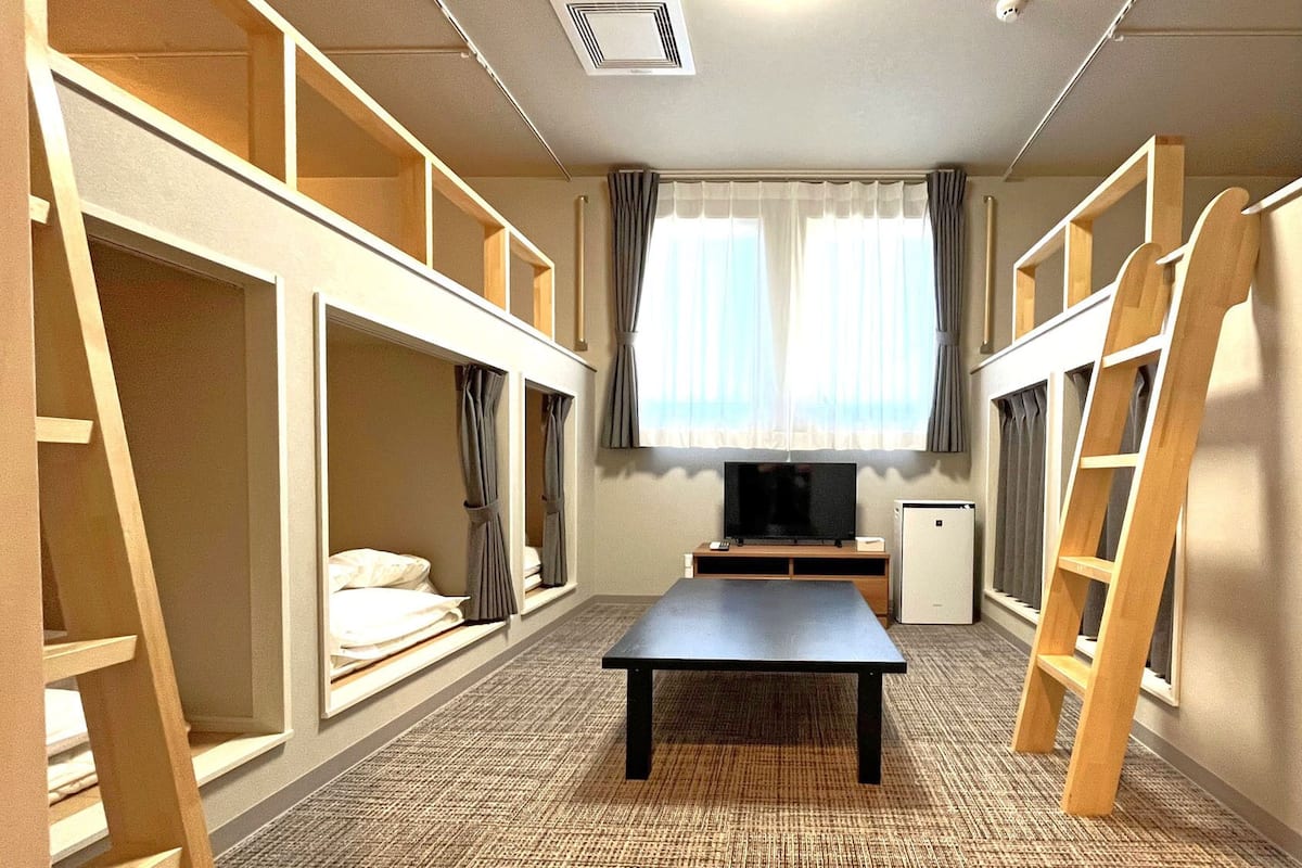 Quarto com beliches, televisão, uma mesa pequena e janela com cortinas.