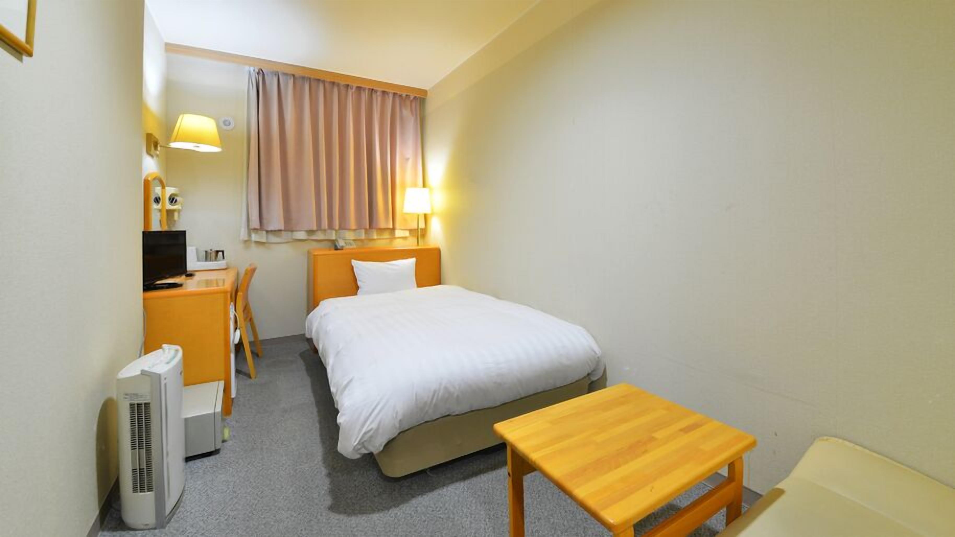Quarto de hotel com uma cama, uma escrivaninha com computador, uma cadeira, uma mesa e uma janela com cortinas.
