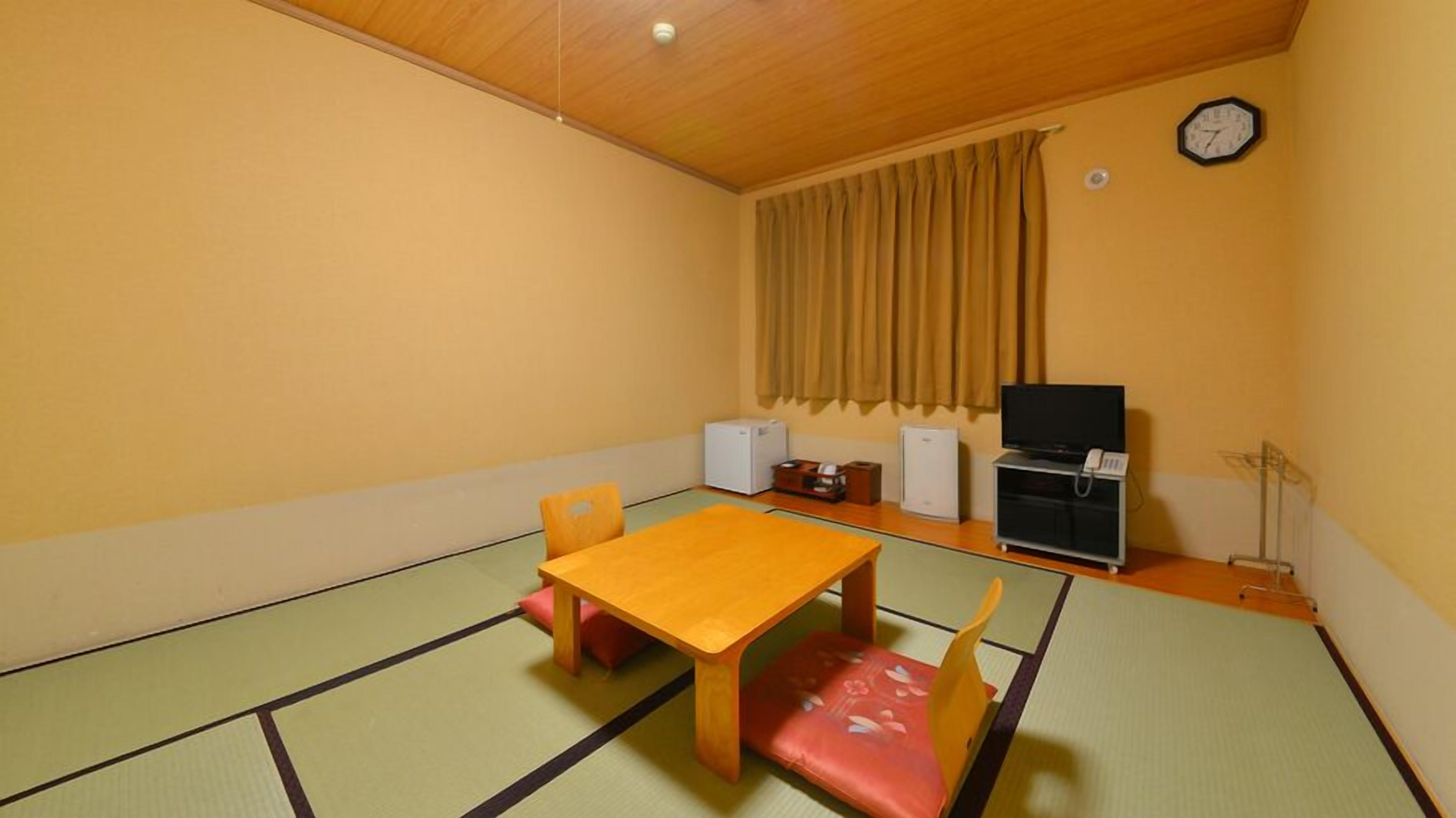 Quarto com tatame, mesa de madeira, cadeiras, televisão, relógio e um pequeno frigorífico.