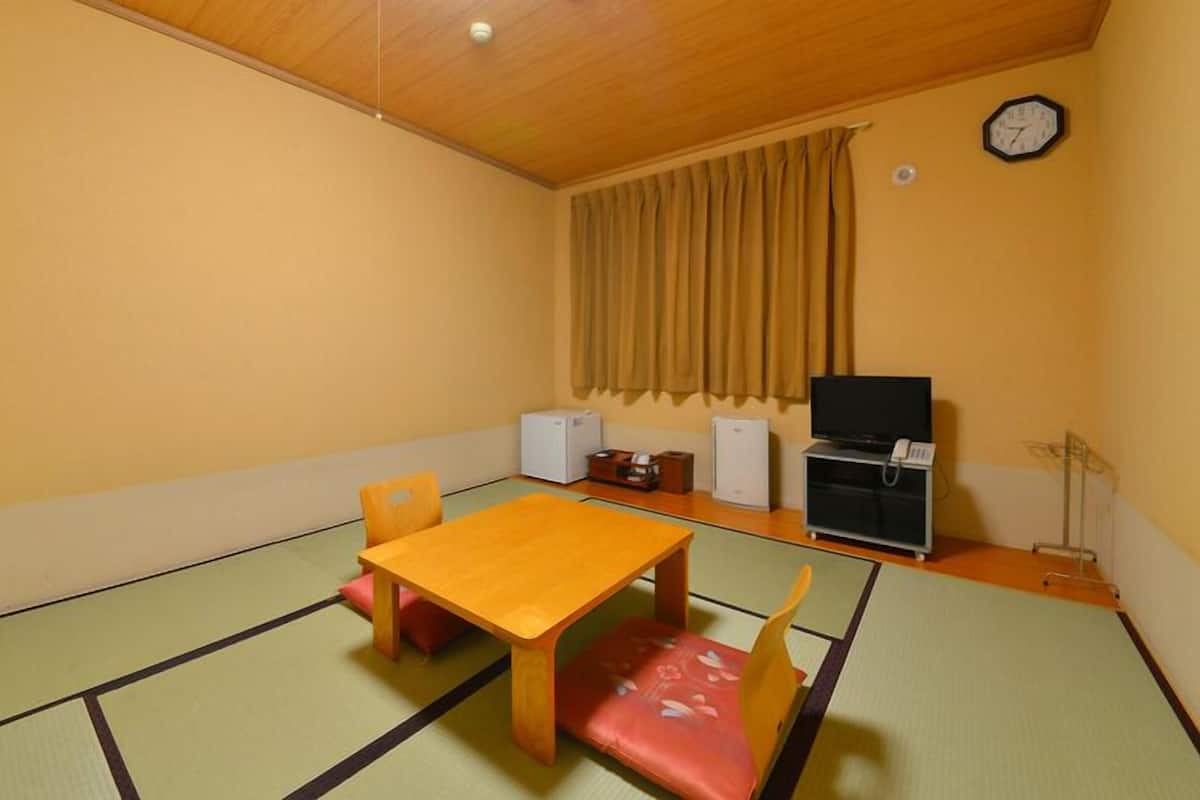 Quarto com tatame, mesa de madeira, cadeiras, televisão, relógio e um pequeno frigorífico.