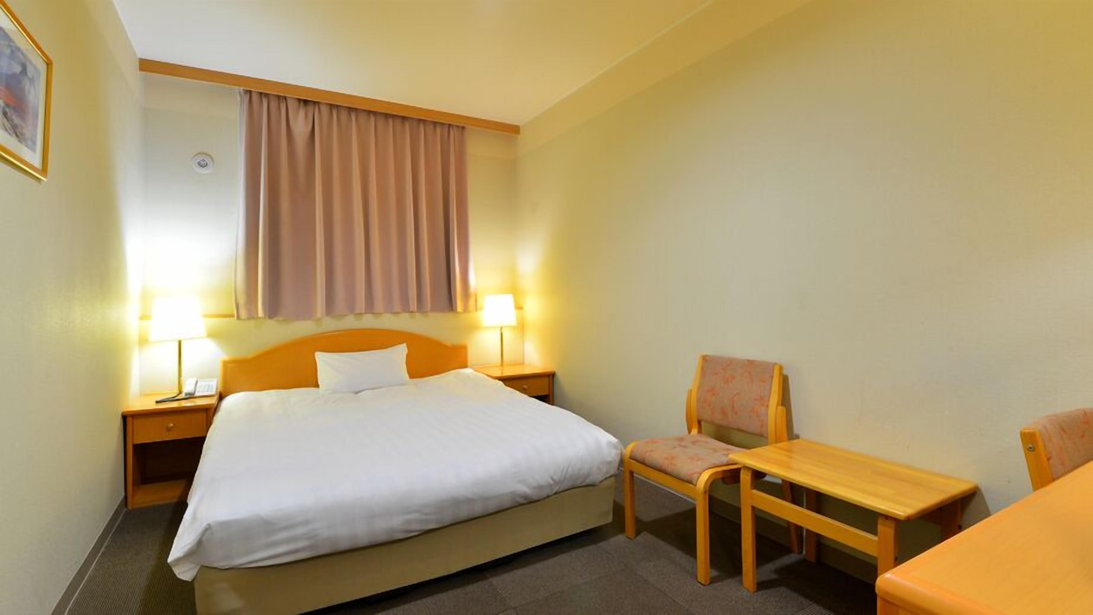 Quarto de hotel com cama, luminárias de cabeceira, uma cadeira e uma mesinha.