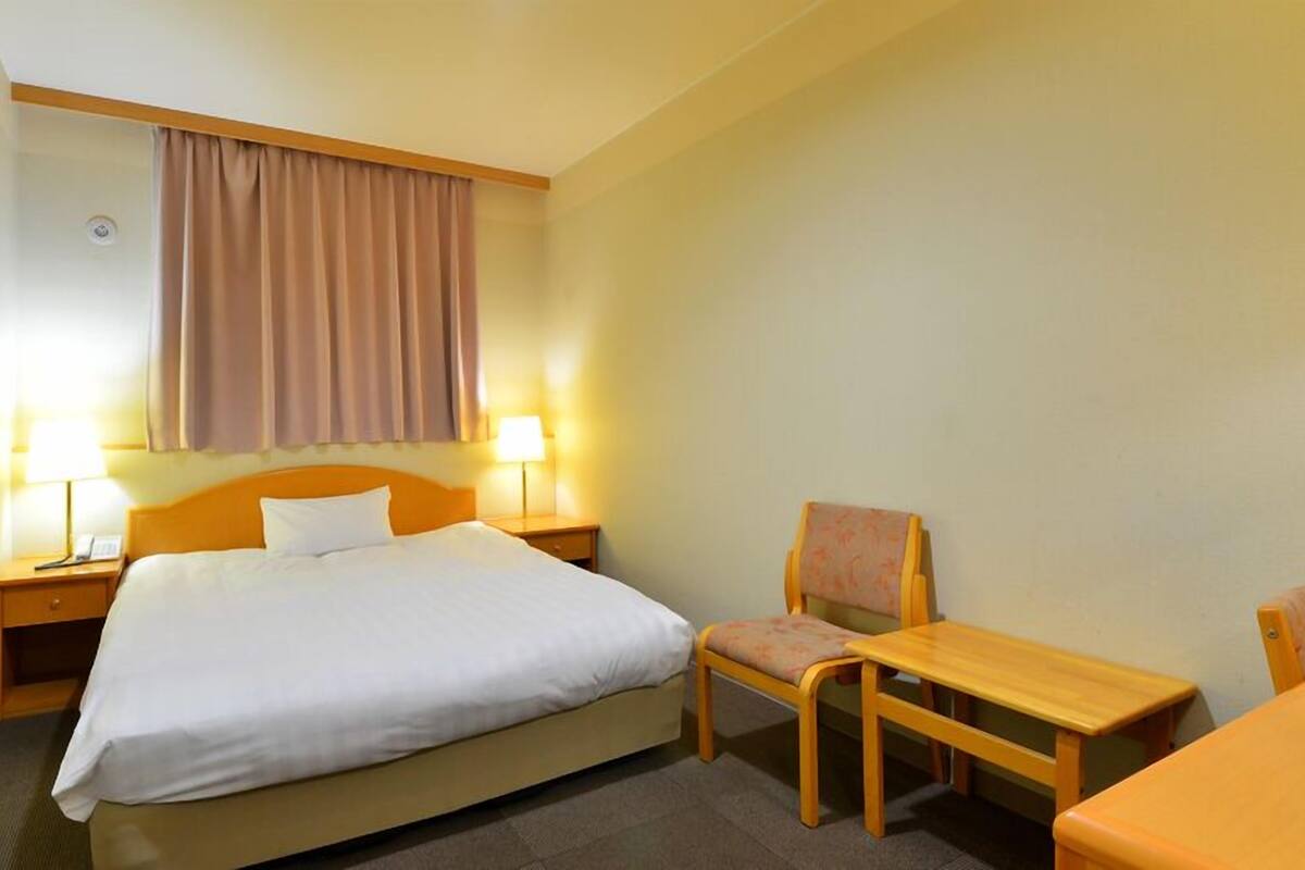 Quarto de hotel com cama, luminárias de cabeceira, uma cadeira e uma mesinha.