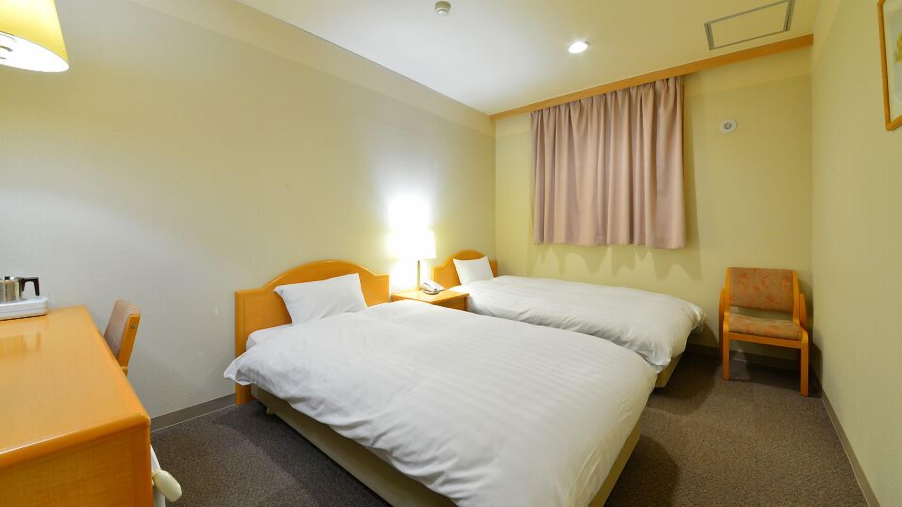 Quarto de hotel com duas camas, uma escrivaninha, uma cadeira, um abajur e uma janela com cortinas.