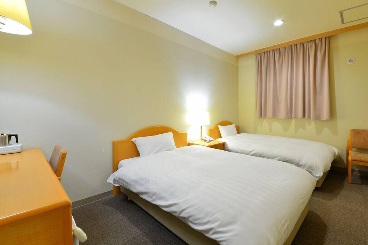 Quarto de hotel com duas camas, uma escrivaninha, uma cadeira, um abajur e uma janela com cortinas.