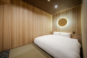 Quarto de hotel moderno com parede revestida em madeira, luminária redonda na parede, cama bem arrumada e um pequeno vaso com flores na mesa de cabeceira.