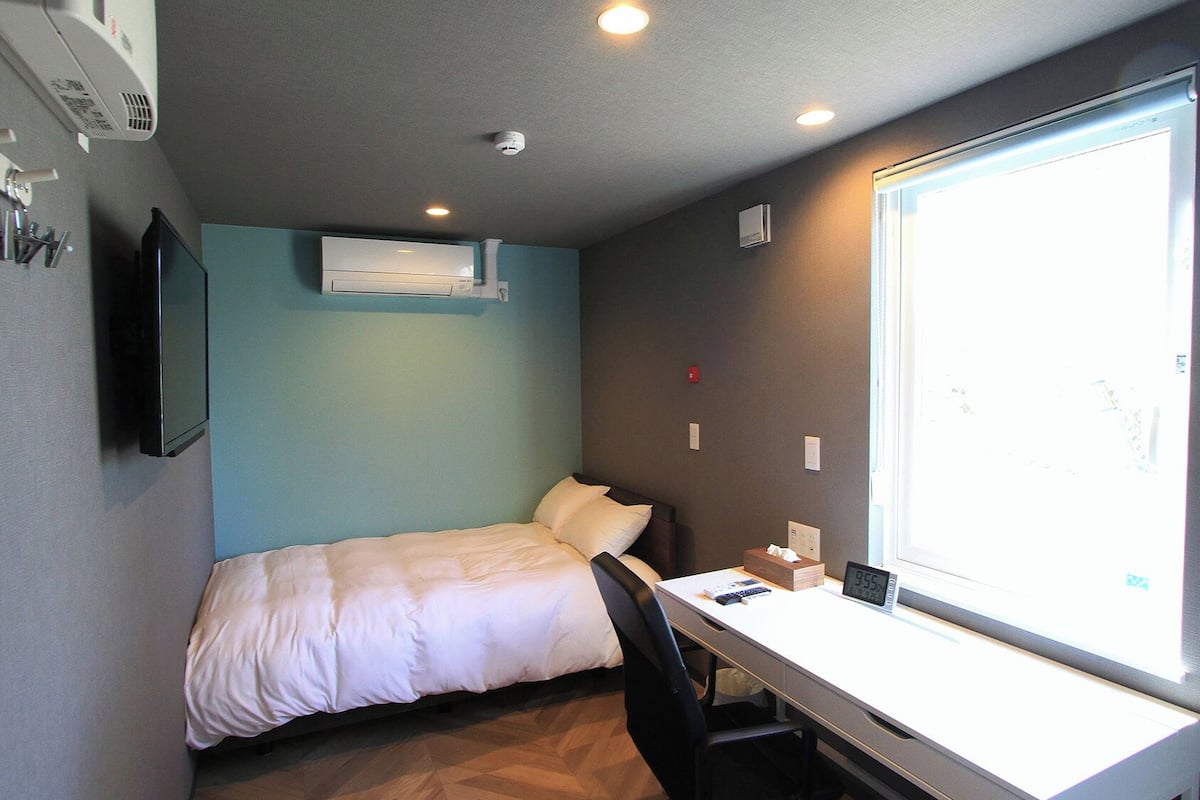 Quarto de hotel compacto com cama, escrivaninha, cadeira e uma janela grande.