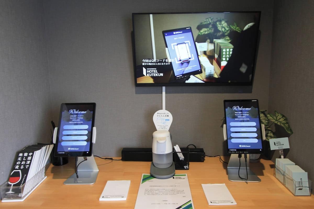 Uma mesa de recepção com dois tablets Lenovo exibindo uma tela de boas-vindas, uma máquina de café e um telefone.