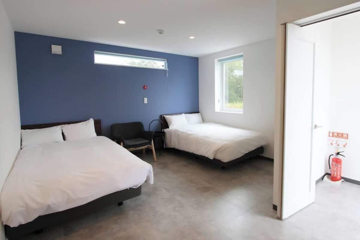Quarto de hotel com duas camas, uma cadeira, uma janela e um extintor de incêndio.