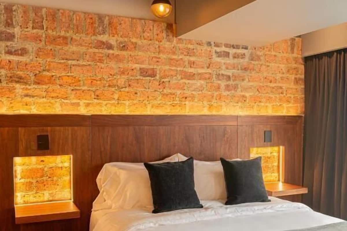 Quarto com parede de tijolos, cabeceira de madeira, cama com roupa de cama branca, duas mesas de cabeceira e iluminação pendente.