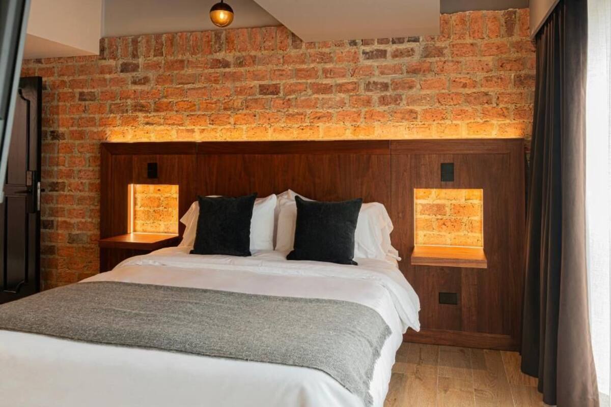 Quarto de hotel com parede de tijolos, cabeceira de madeira, duas luminárias de apoio, cama com roupa de cama branca e manta cinza.