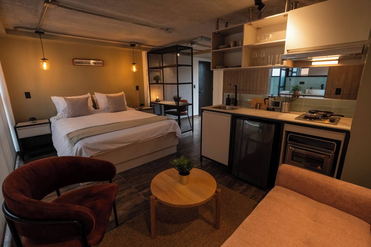 Quarto de hotel compacto com cama, pequena mesa de jantar, cadeira, cozinha compacta e área de estar separada com sofá.