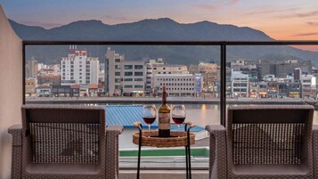 Quarto com vista para um horizonte urbano e montanhas, duas cadeiras de vime, uma mesa redonda pequena com uma garrafa e copos.