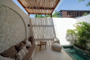 Uma área de estar moderna ao ar livre, com muro de pedra, pérgola de madeira, um banquinho pequeno de madeira e um elemento decorativo com água.