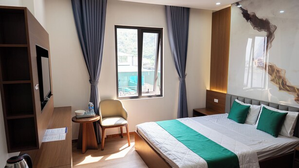 Quarto de hotel com cama, cadeira, mesa pequena, chaleira e janela com cortinas.