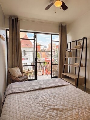 Quarto com cama, ventilador de teto, estante e vista da cidade por grandes janelas.