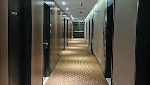 Um corredor comprido e estreito com carpete estampado, portas em ambos os lados e iluminação embutida no teto.