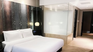 Quarto de hotel moderno com uma cama grande, divisória de vidro e uma área de estar separada.