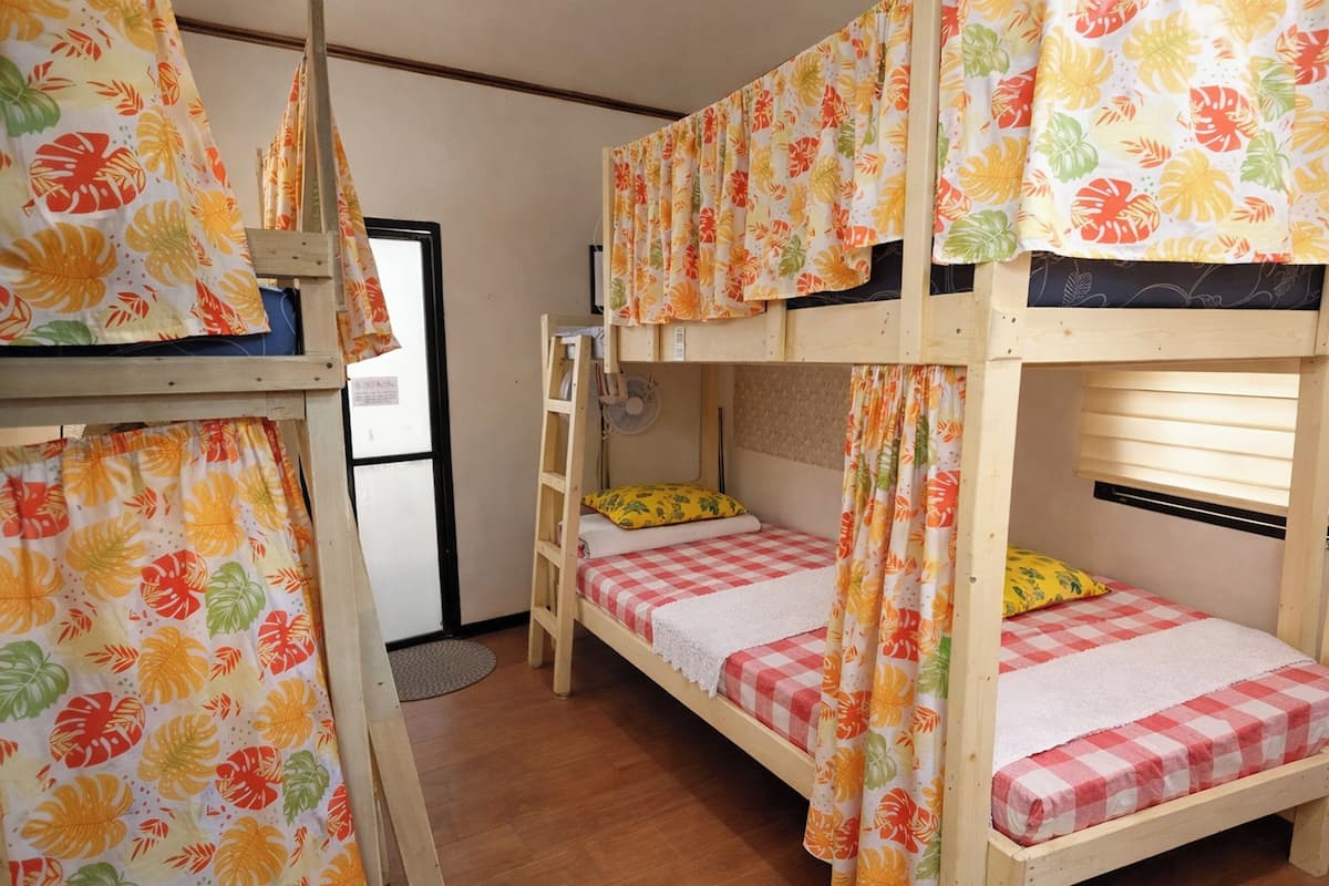 Quarto com beliche, cortinas florais, ventilador e colcha xadrez.