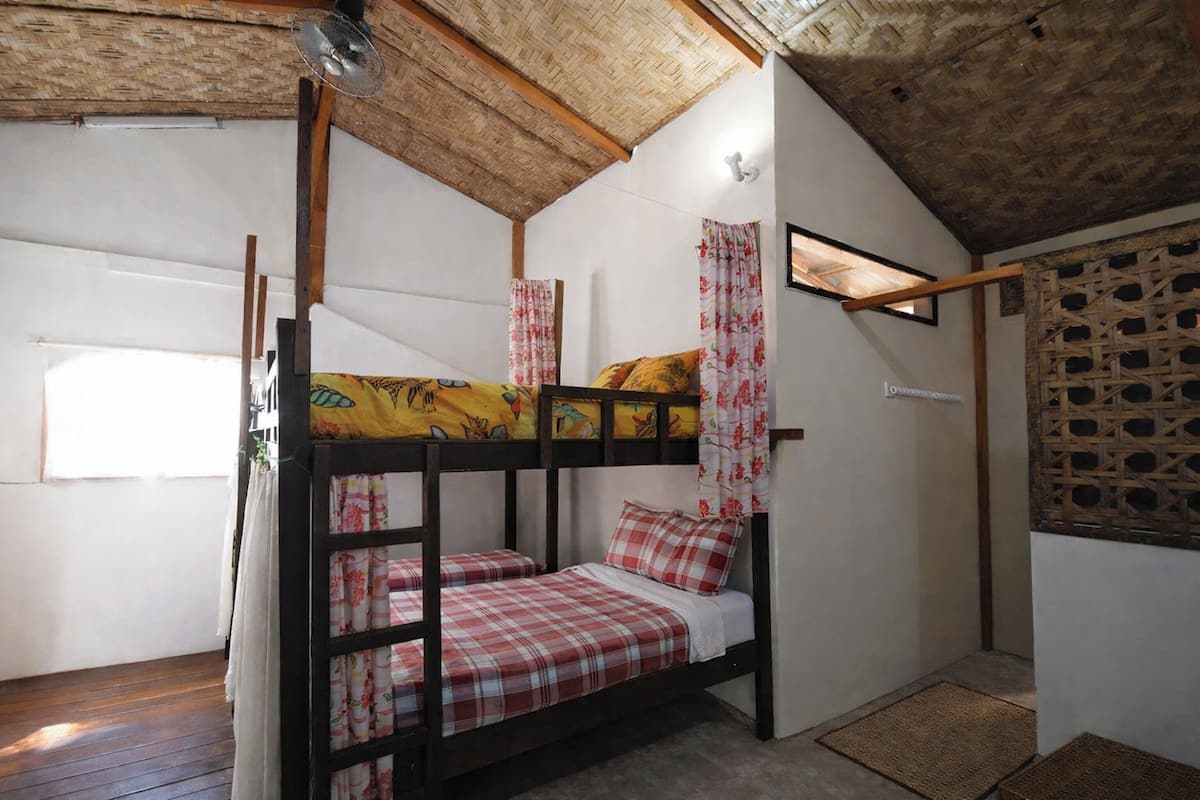 Quarto com beliche, teto de palha, uma pequena janela e luminária fixa na parede.