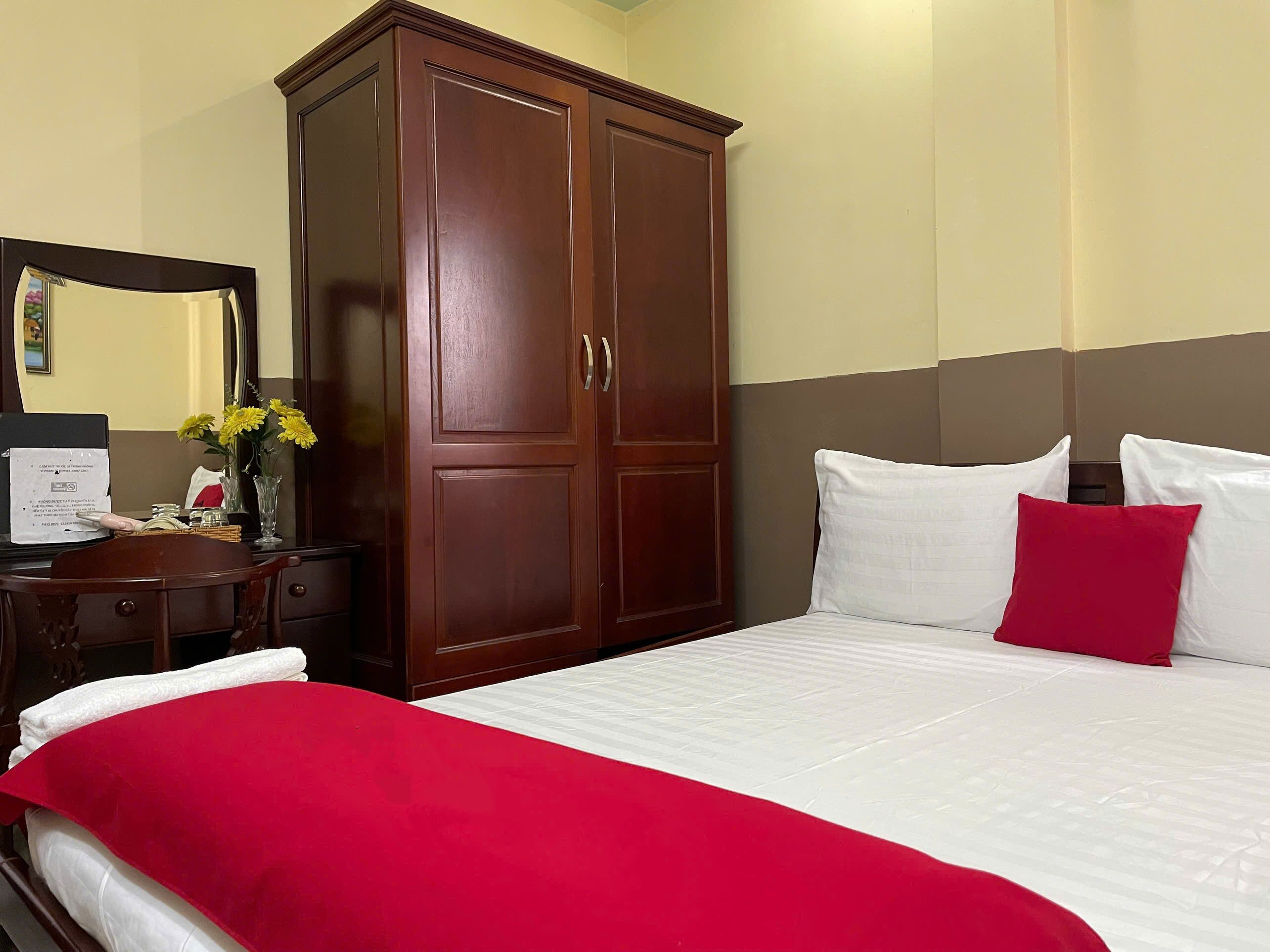 Quarto de hotel com cama, guarda-roupa, escrivaninha com espelho e um vaso com flores.