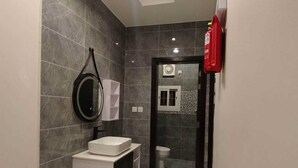 Um banheiro moderno com toucador branco, paredes revestidas de azulejos escuros, espelho redondo e extintor de incêndio vermelho.
