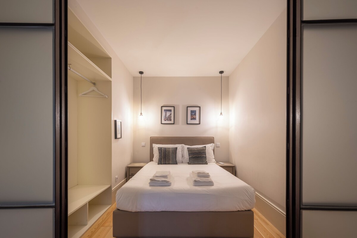 Quarto com cama, mesas de cabeceira, guarda-roupa e duas luzes fixadas na parede.