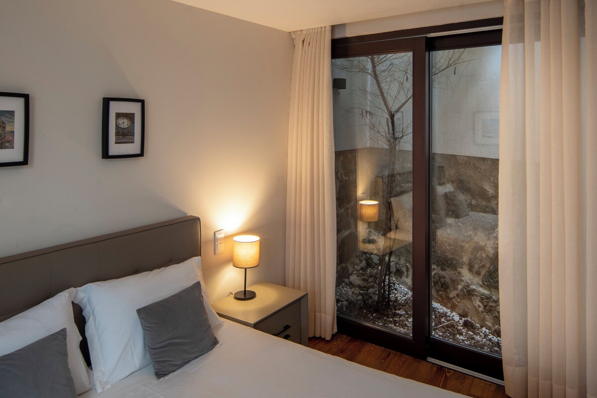 Quarto de hotel com uma cama, duas almofadas, uma mesa de cabeceira com abajur e uma janela com cortinas.