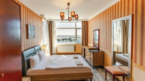 Quarto de hotel com uma cama grande, cômoda, um banquinho, um lustre e vista para o exterior.