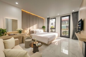 Quarto de hotel moderno com cama, sofá, mesas de cabeceira, televisão e varanda com cortinas.