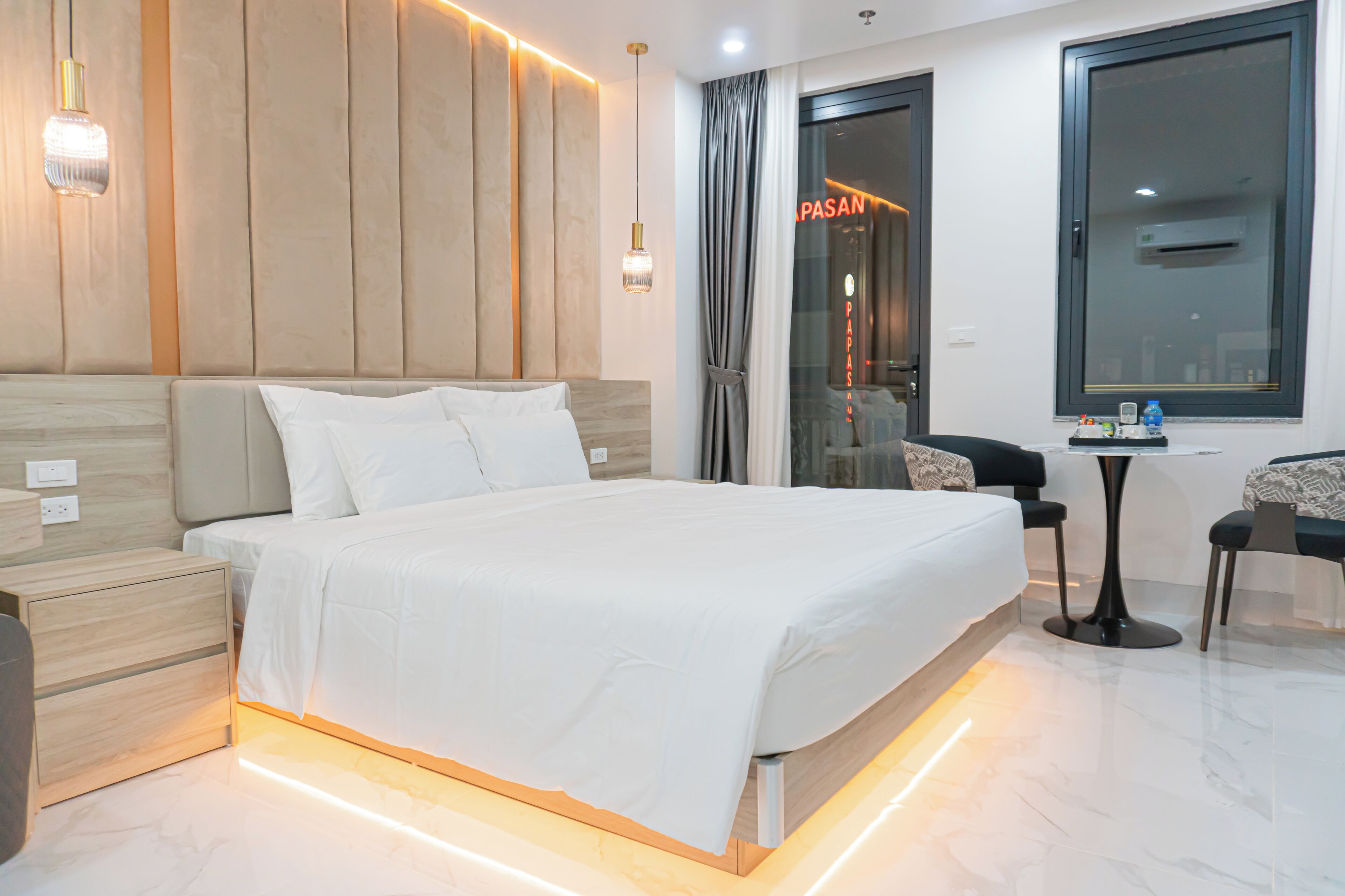 Quarto de hotel moderno com uma cama grande, mesas de cabeceira, uma pequena mesa com cadeiras e vista para a rua.