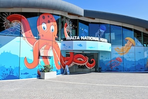 Um edifício com um grande mural de polvo, o nome 'MALTA NATIONAL AQUARIUM' na fachada e uma área de entrada com azulejos.