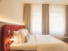 Quarto de hotel com uma cama, mesa de cabeceira, abajur e janela com cortinas.