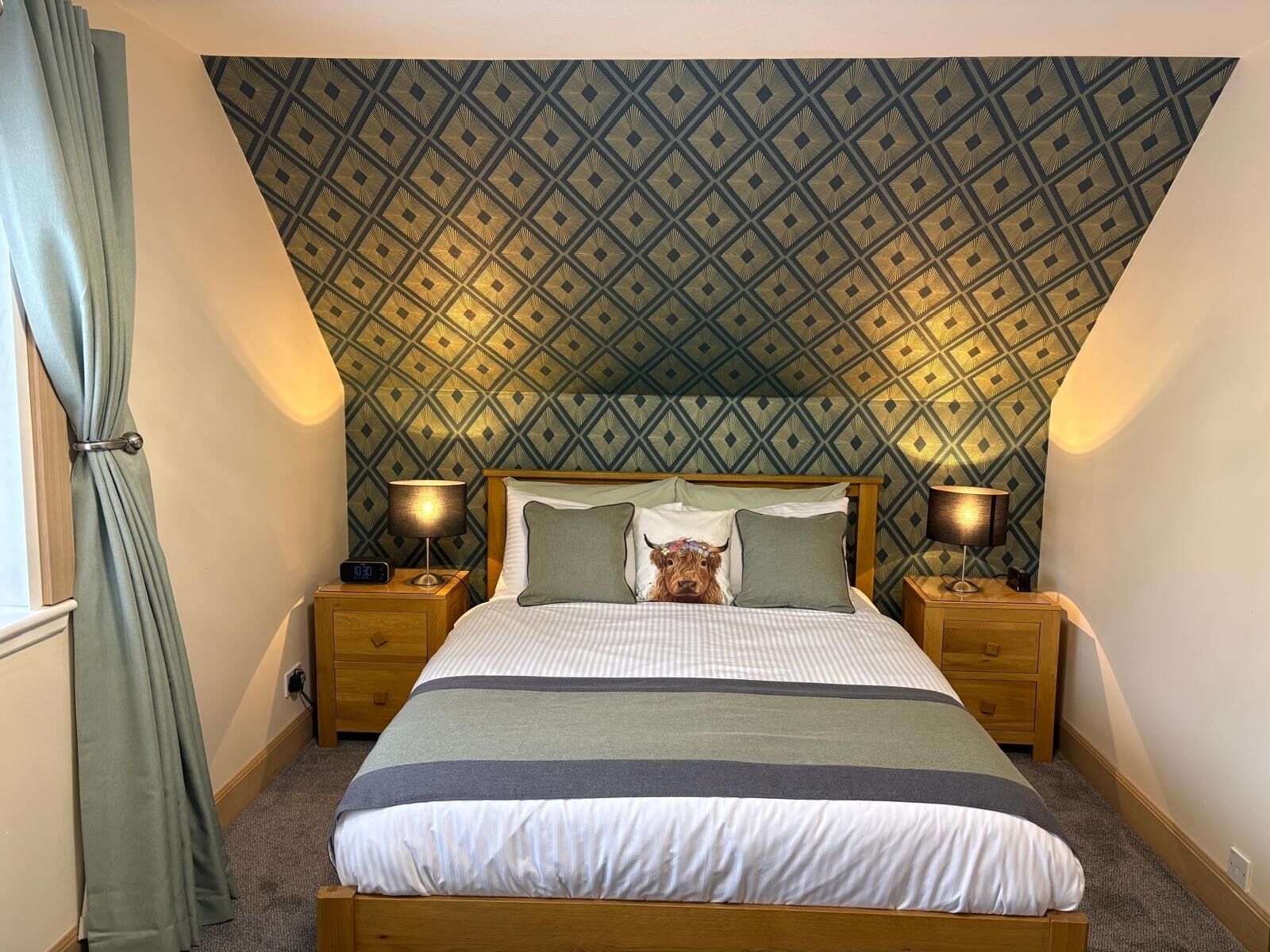 Um quarto com papel de parede estampado, uma cama com colcha listrada, duas mesas de cabeceira com luminárias e uma janela com cortinas.