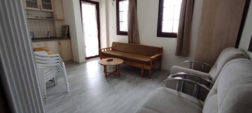 Uma sala de estar com sofá, mesa redonda, uma cadeira e cozinha com armários e janela.