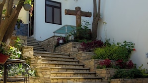 Uma escadaria de pedra leva a um edifício branco com uma cruz, rodeado por vegetação e uma palmeira.