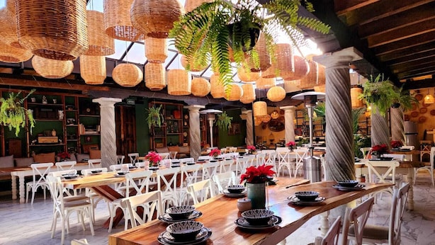 Um restaurante com mesas de madeira compridas, cadeiras brancas, luminárias de vime pendentes e plantas em vasos.