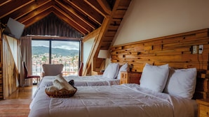 Quarto com duas camas, cabeceira de madeira, janela com vista para uma cidade e uma cadeira vermelha.