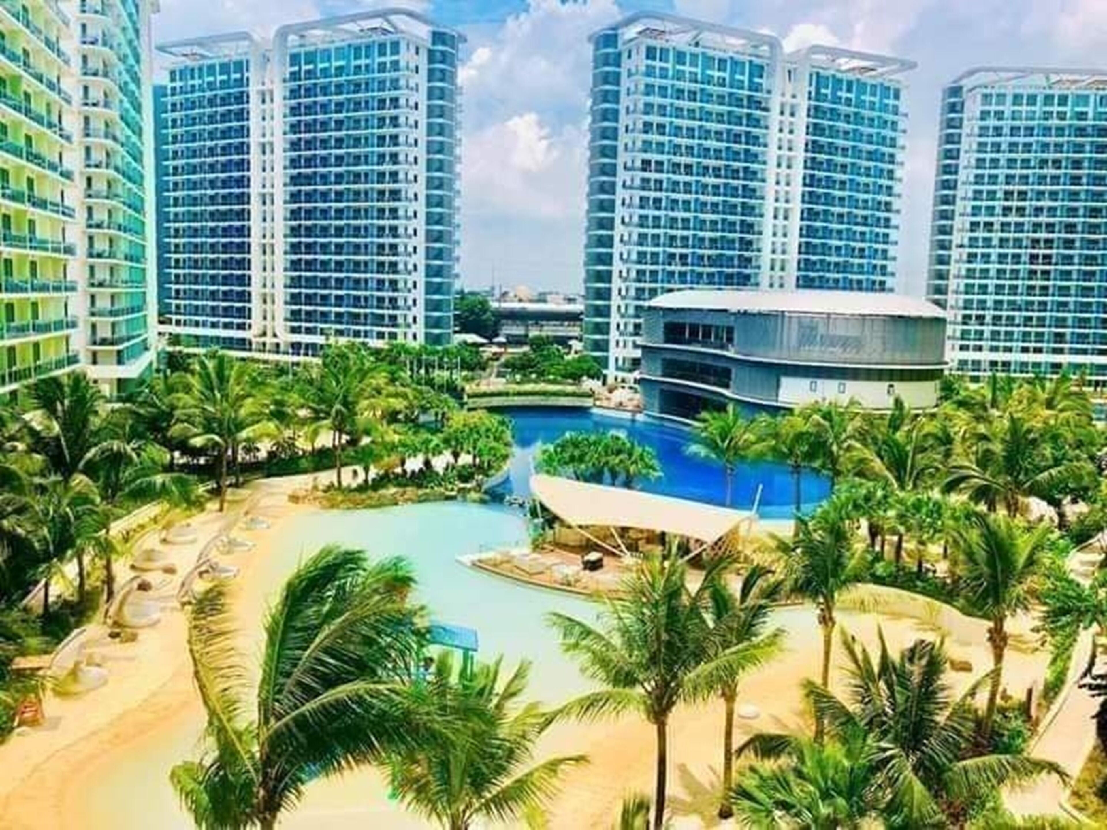 Um hotel alto com uma piscina central cercada por palmeiras e céu claro.